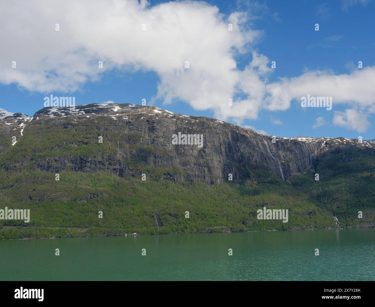 A steep green cliff rises above calm water, surrounded by a clear blue ...