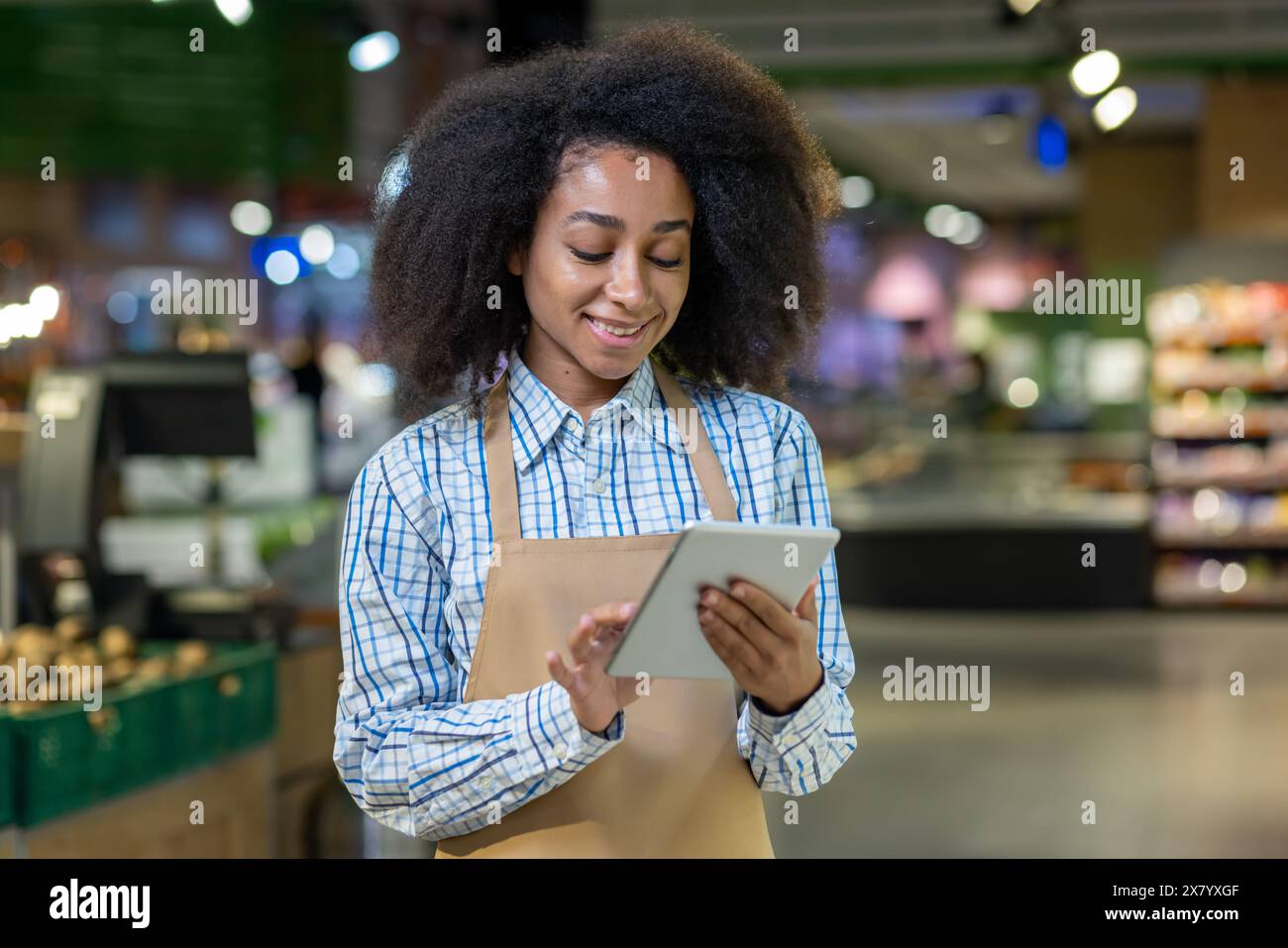 An employee with afro hair, wearing an apron, uses a tablet in a ...