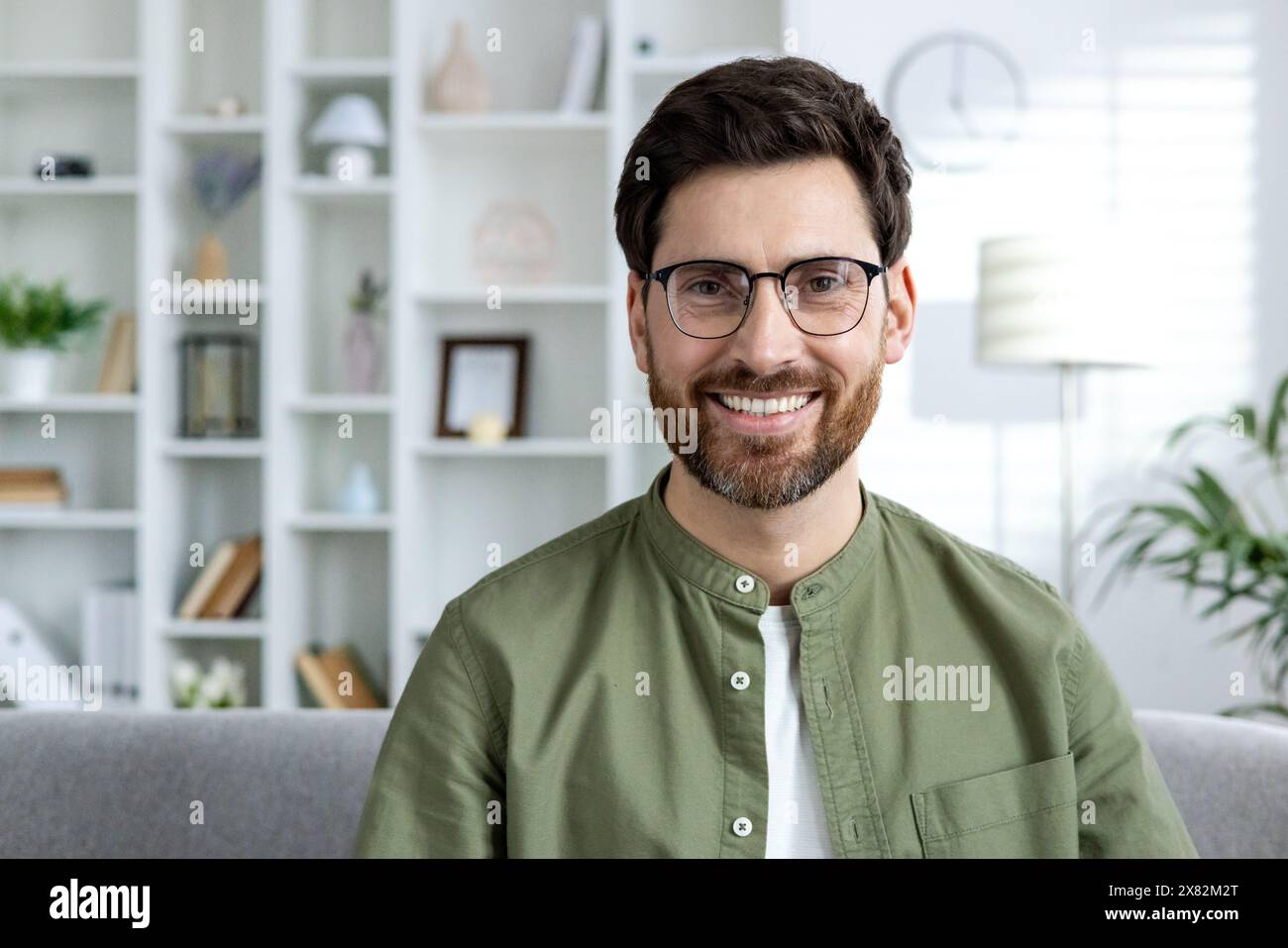 Casually dressed man with glasses smiling at home, sitting on a couch ...