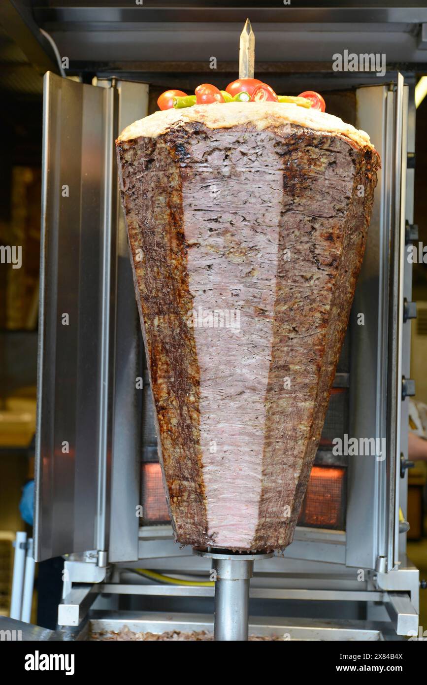 Large piece of kebab meat on a spit, topped with tomatoes, in a fast ...