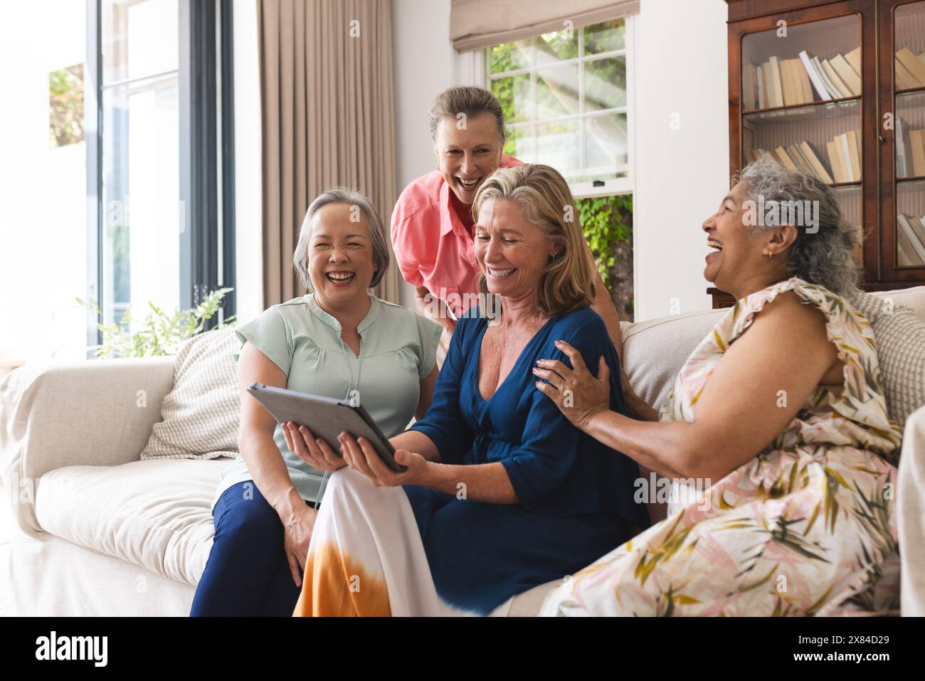 Diverse senior female friends laughing at home, one holding tablet ...