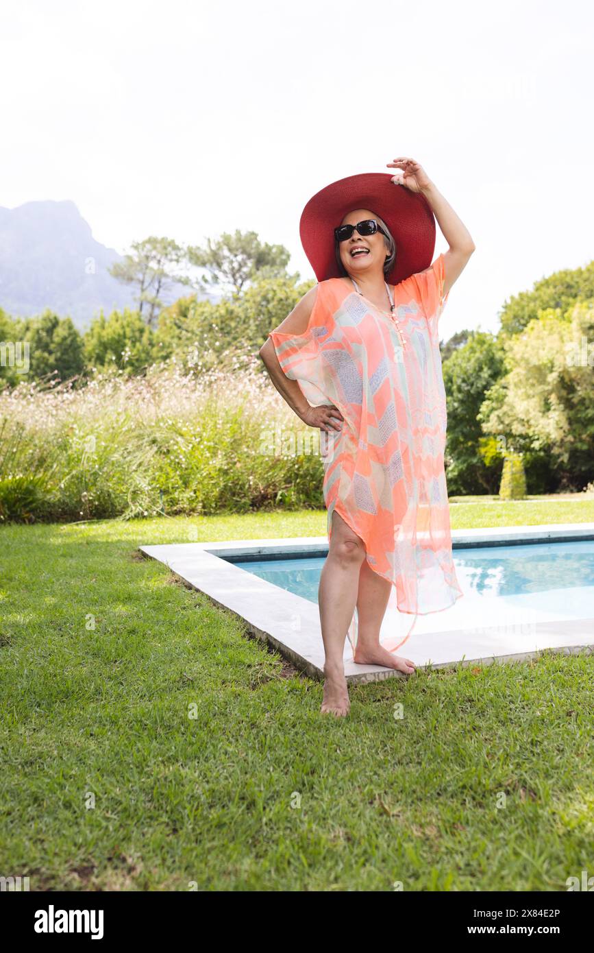 Outdoors, Asian senior female wearing large red hat, standing by a pool ...