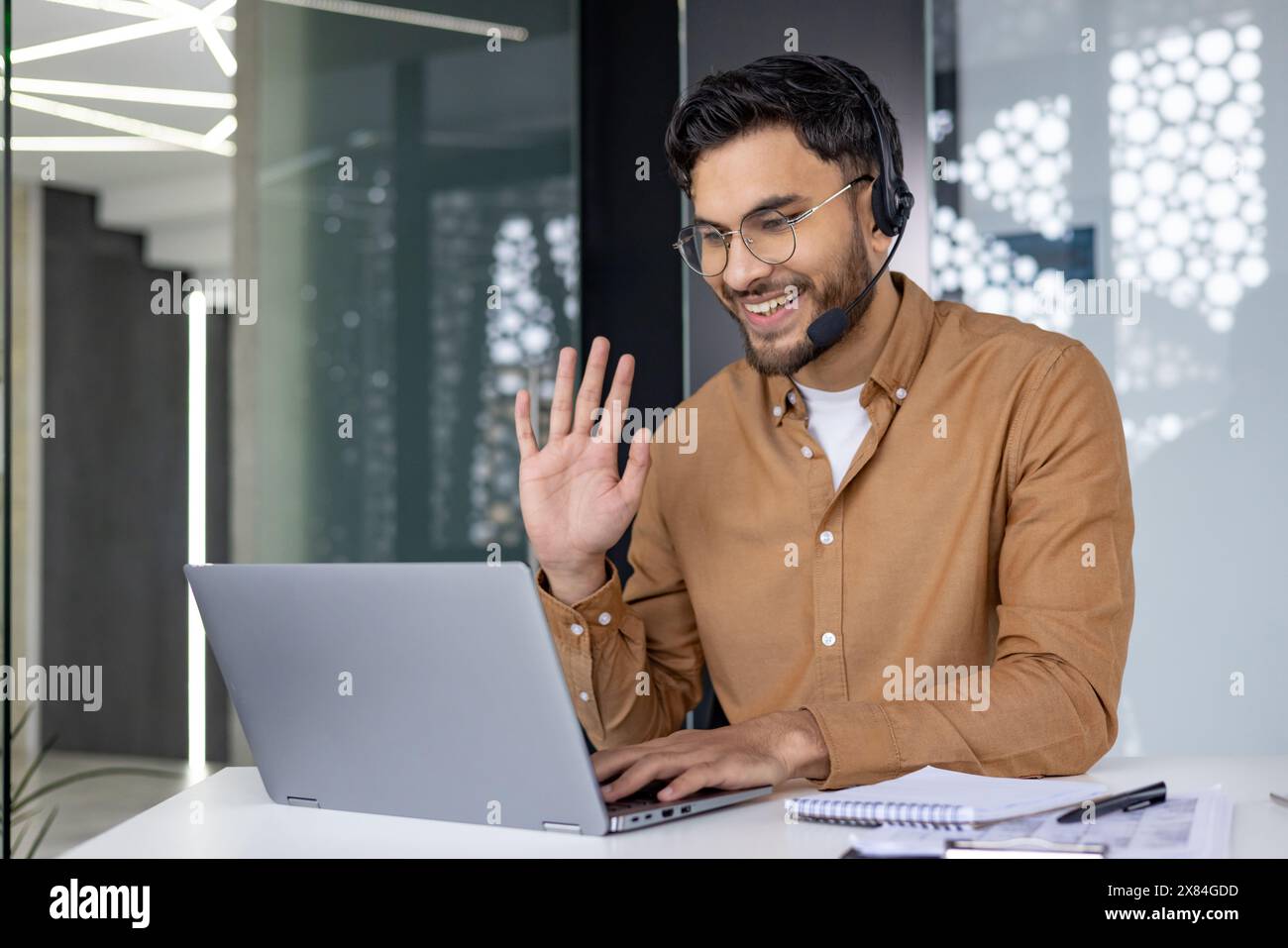A smiling man wearing a headset and waving during an online video call ...