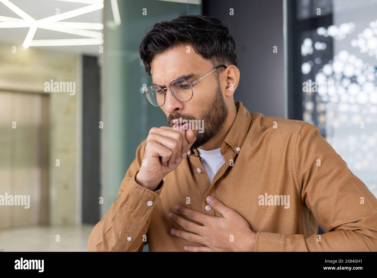 A young man coughing with hand on chest in a modern office environment ...