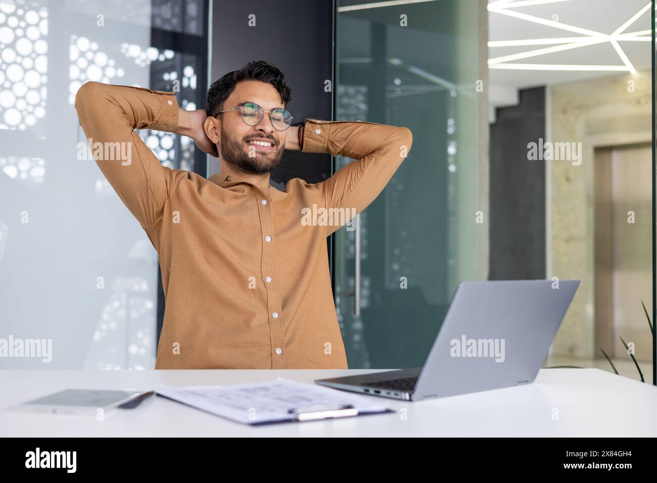 Smiling man relaxing at his desk with hands behind head, taking a break ...