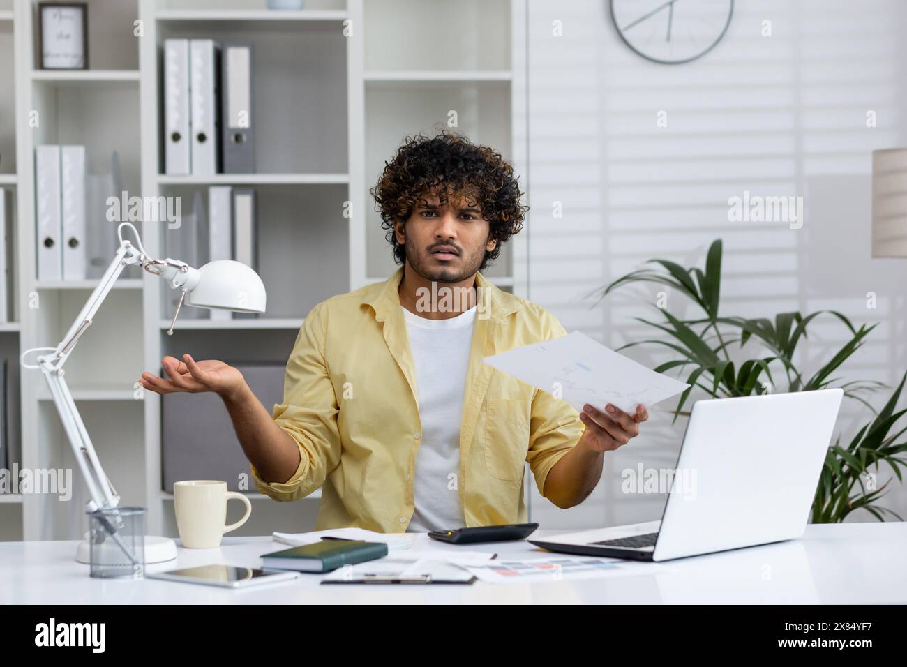 A confused man sitting at his desk in a home office, holding documents ...