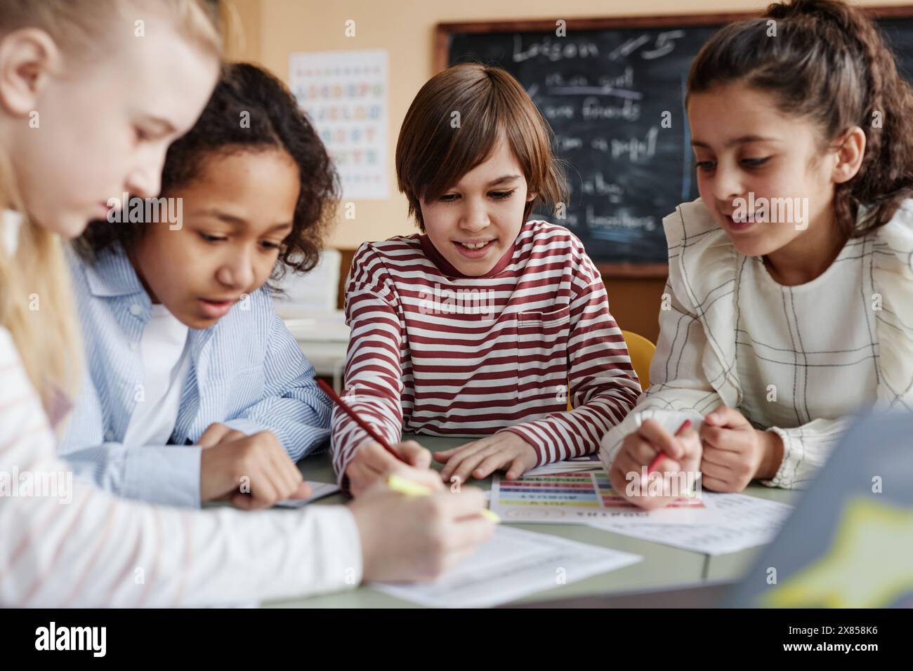 Group of ethnically diverse kids sitting at table in classroom doing ...