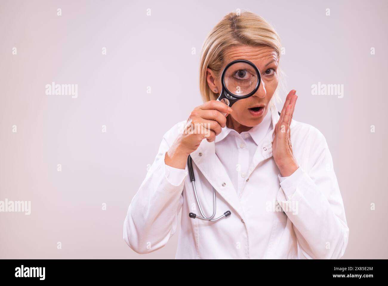 Portrait of shocked mature female doctor with loupe on gray background ...