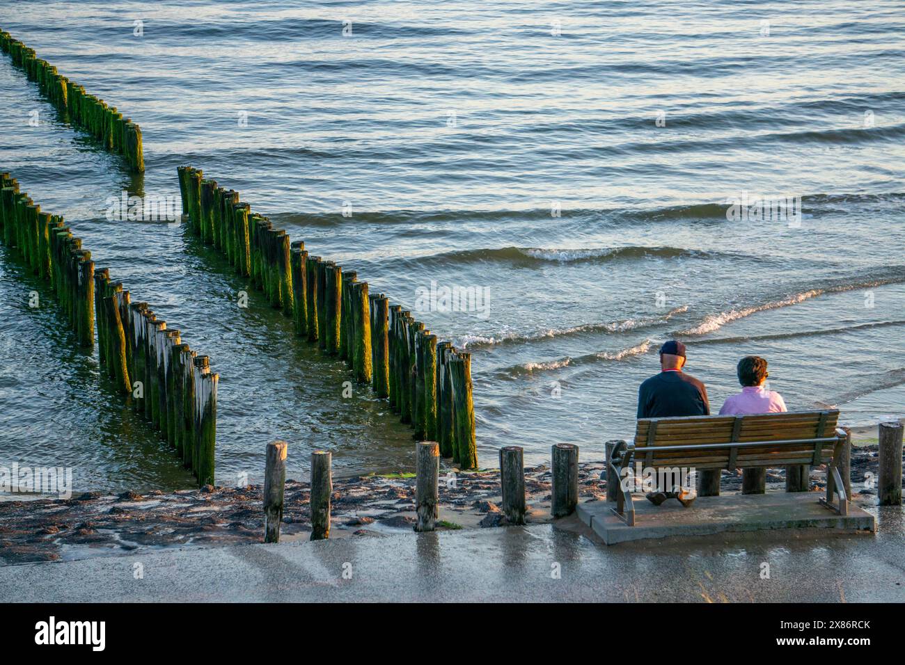 An elderly couple sits on a bench on the beach, at high tide ...