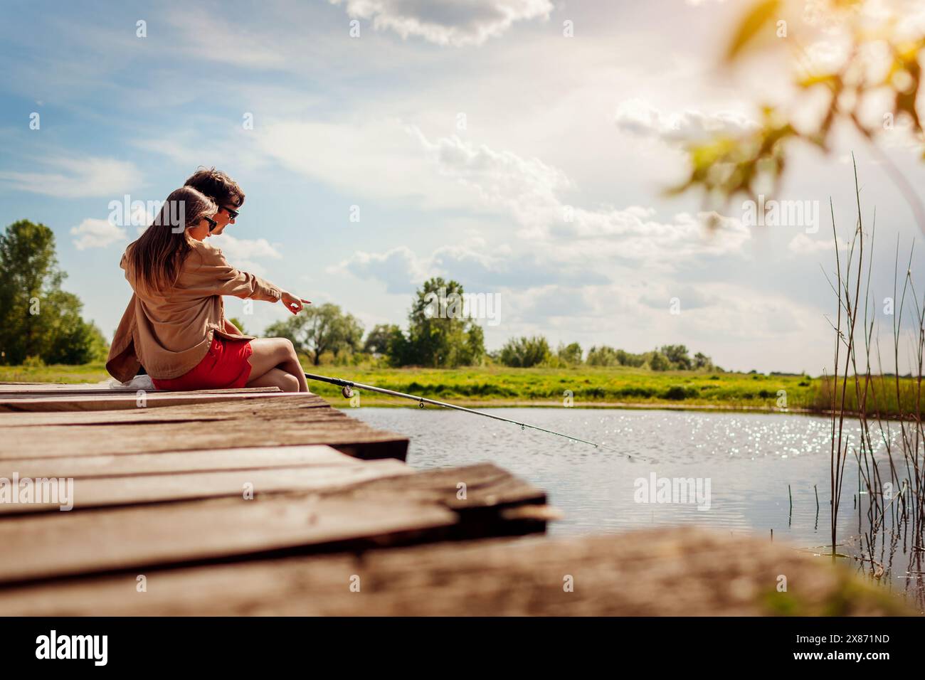 Young couple in love fishing sitting on bridge across lake holding rod ...