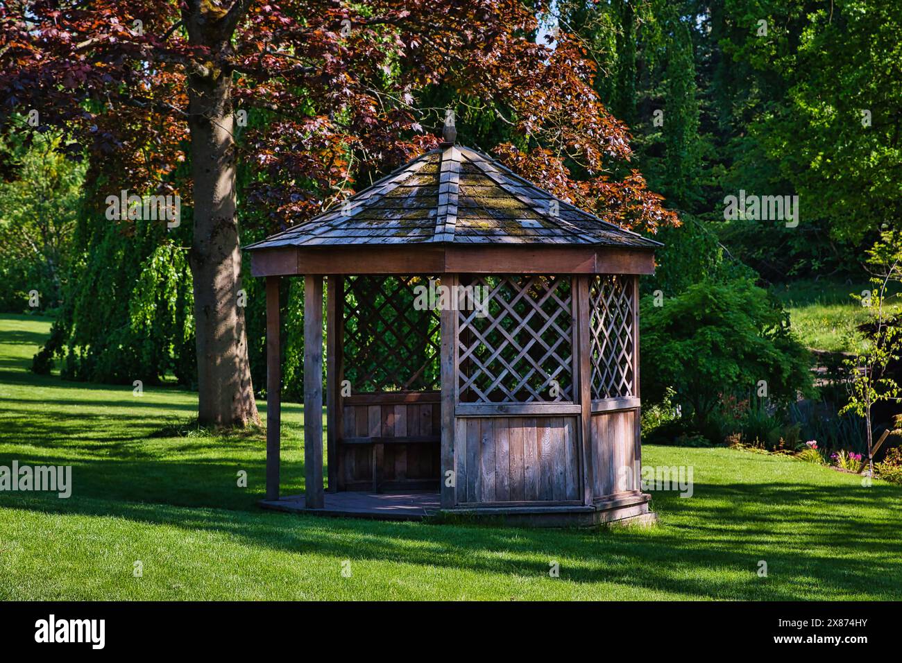 A wooden gazebo with a shingled roof in a lush green garden. The gazebo has lattice walls and is ...