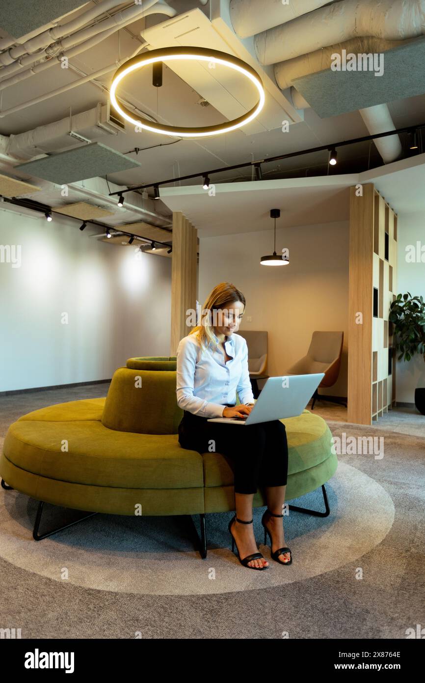 Woman seated on a green circular couch types on her laptop in a modern ...