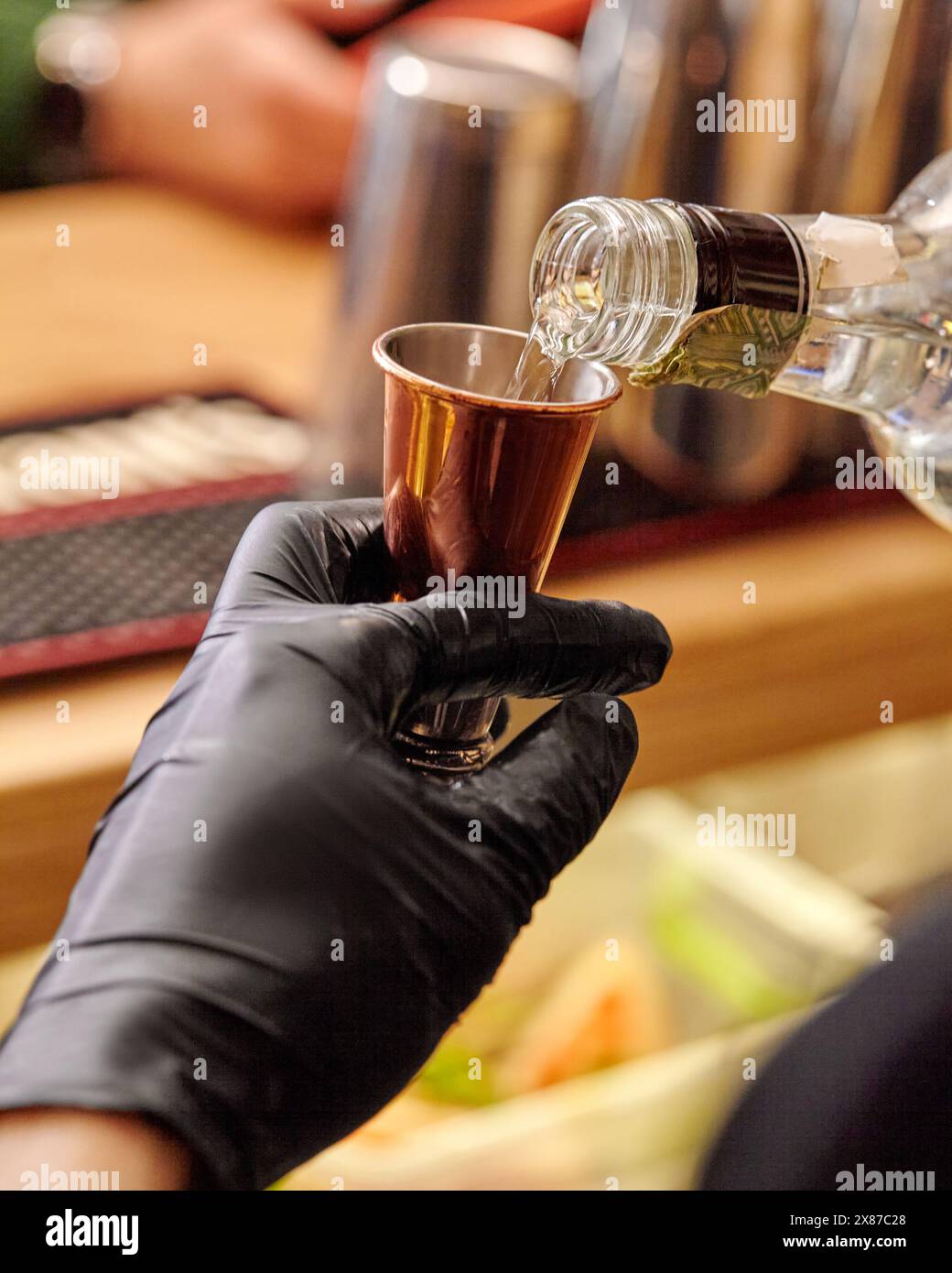 Bartender hand in black glove expertly pouring clear alcohol drink from bottle into gleaming ...