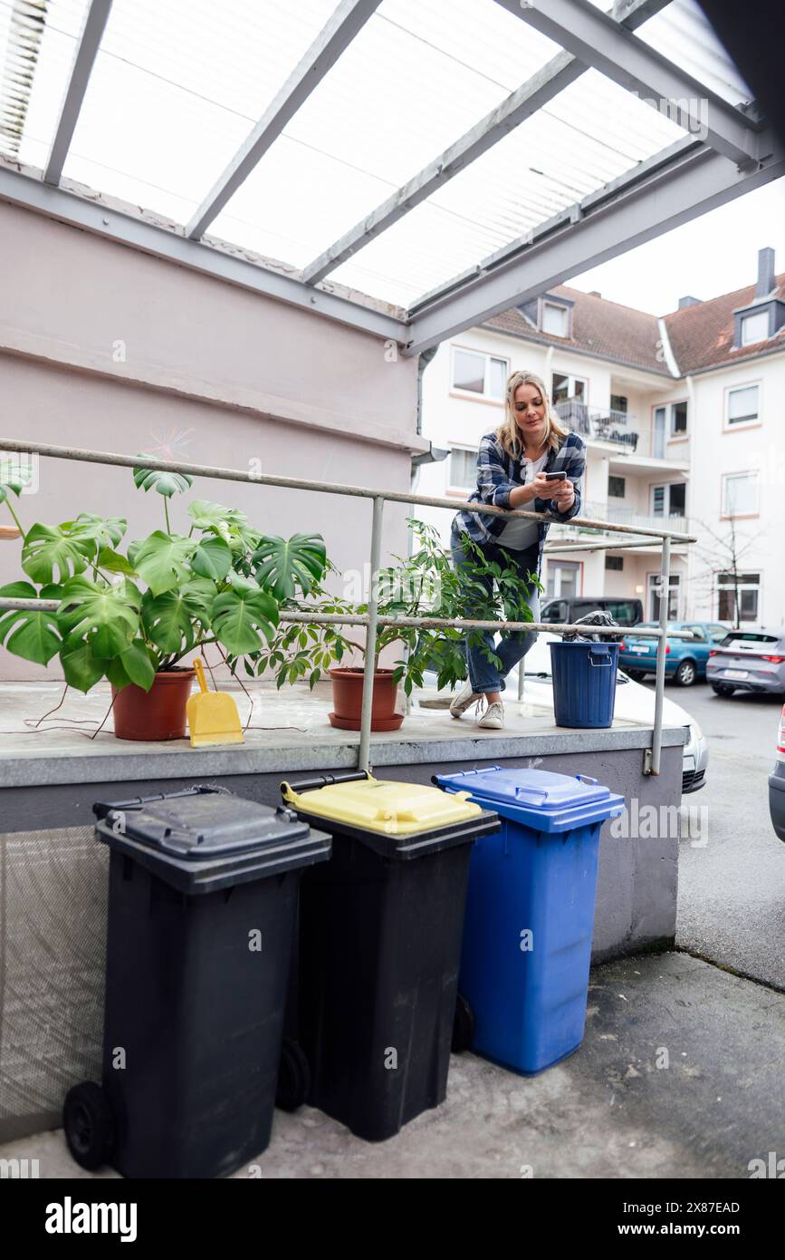 Mature woman leaning on railing using smart phone near garbage bins ...
