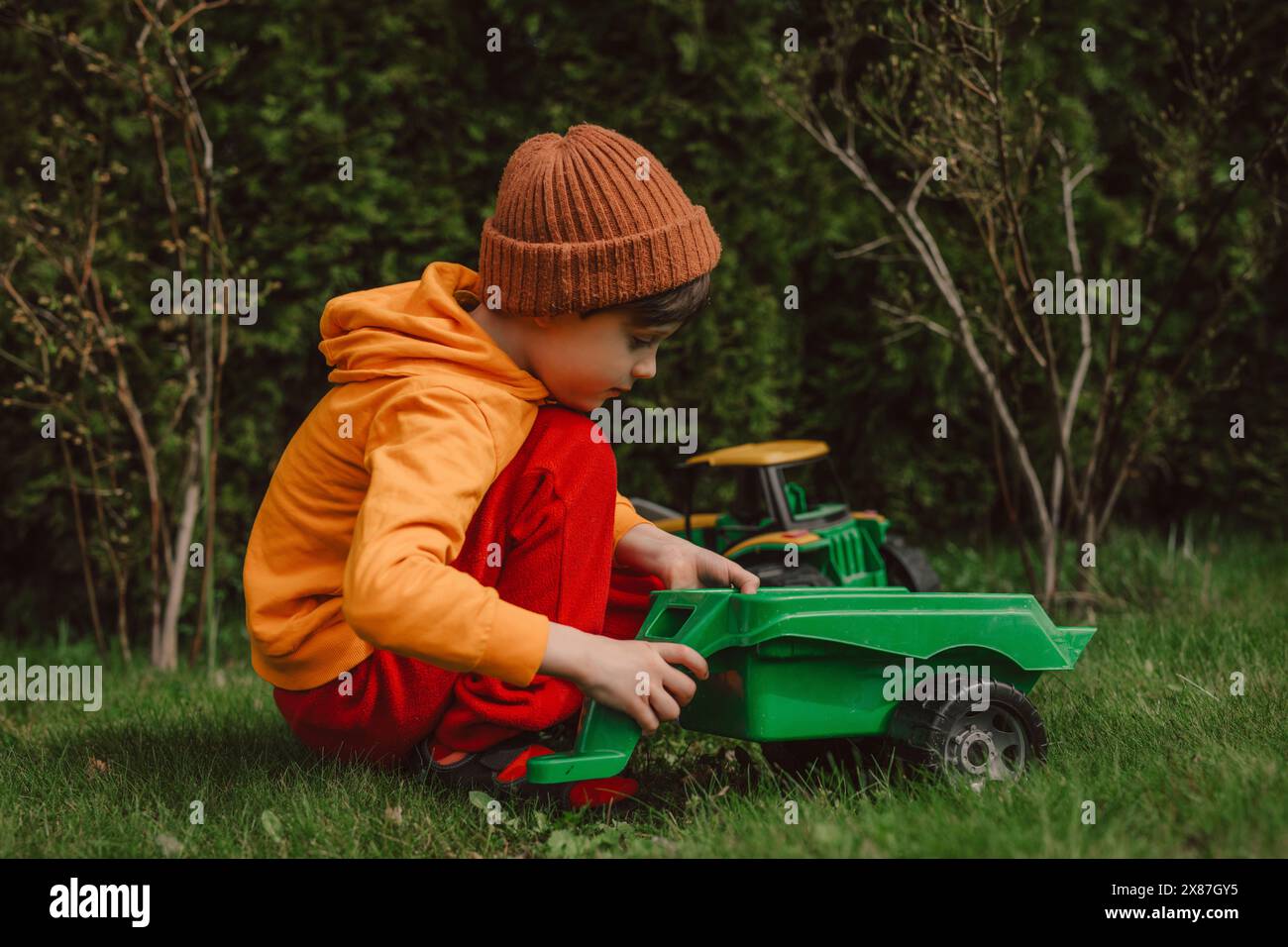 Boy crouching and playing with green toy tractor on grass in back yard ...