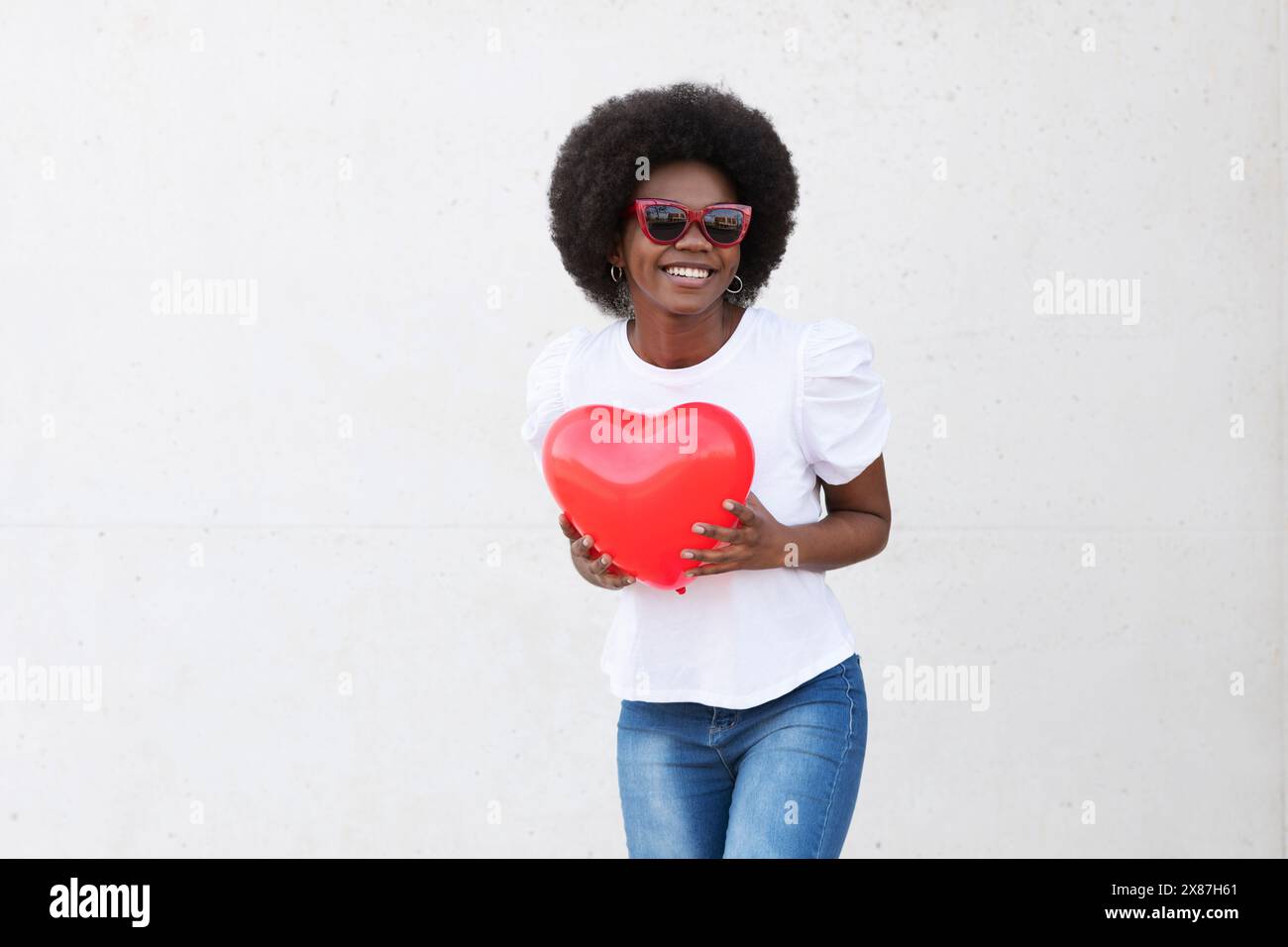 Smiling young Afro woman holding red heart shape balloon standing in ...