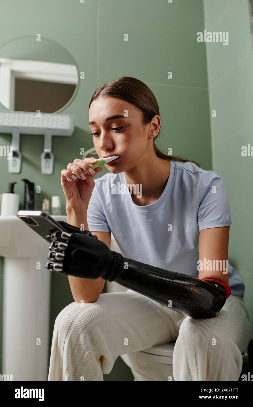 Young woman with black bionic arm brushing teeth and using smart phone ...
