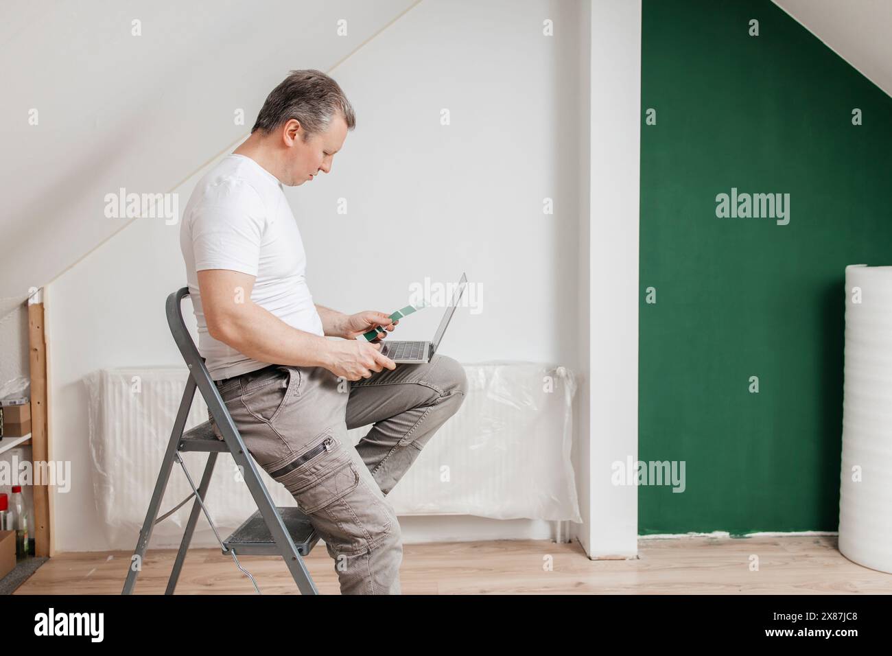 Man holding color swatch and using laptop sitting on stepladder at home ...
