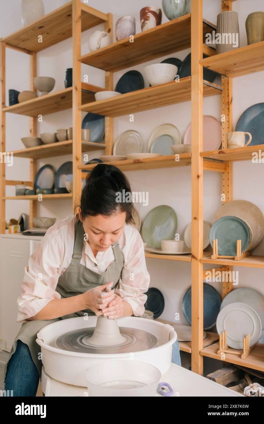 Confident craftsperson molding clay on pottery wheel by rack at ...