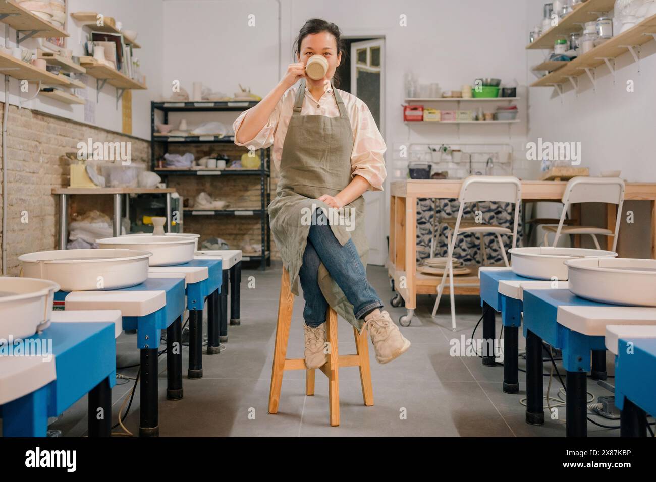 Craftsperson drinking coffee sitting on stool amidst pottery wheels at ...
