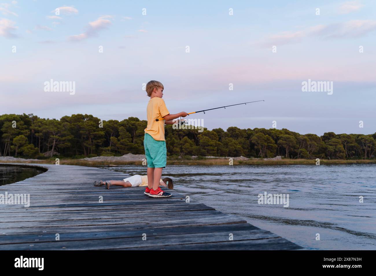 Brother with fishing rod standing by sister lying on pier over lake at ...