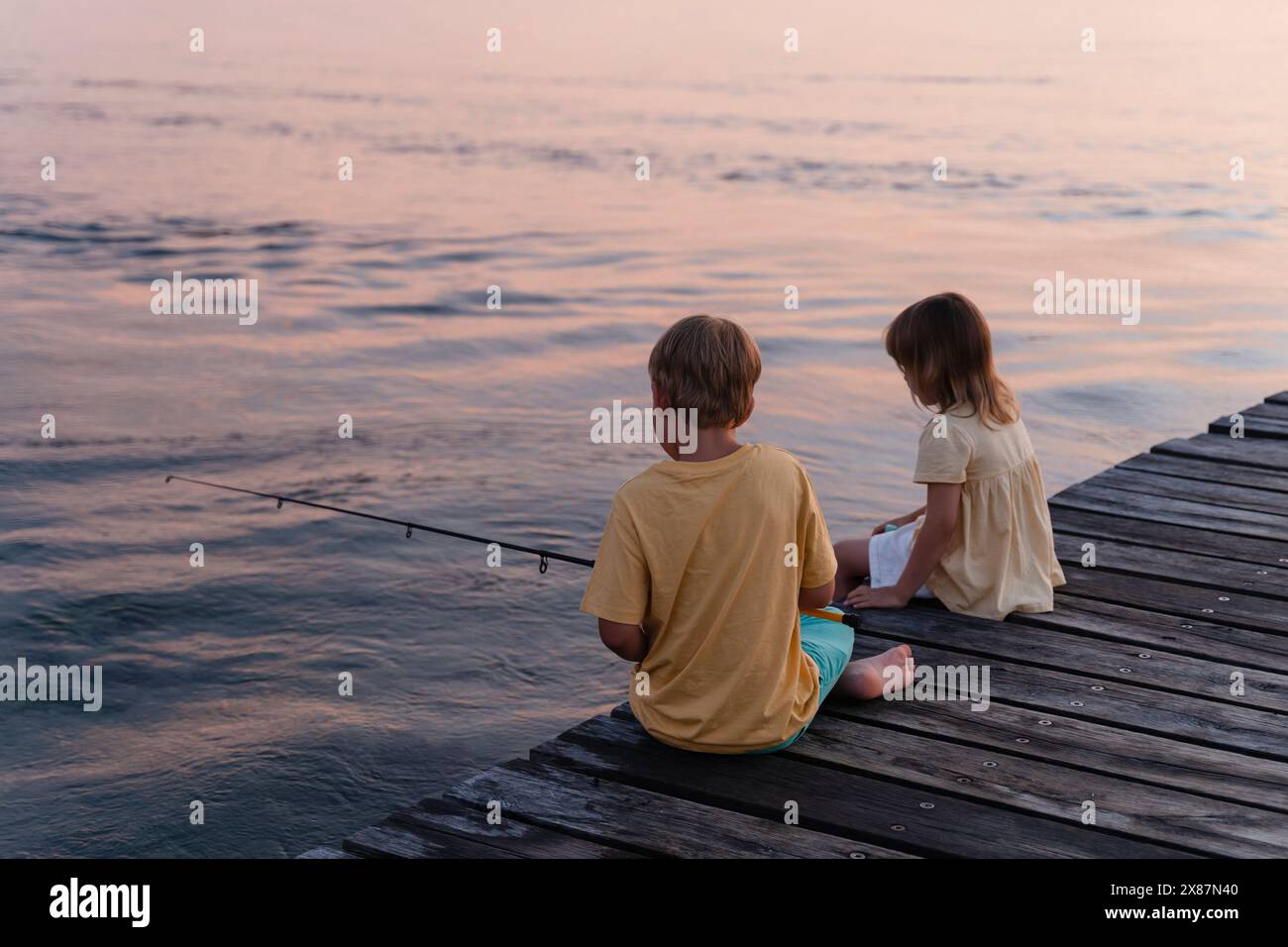 Brother and sister fishing together sitting on pier in lake at sunrise ...
