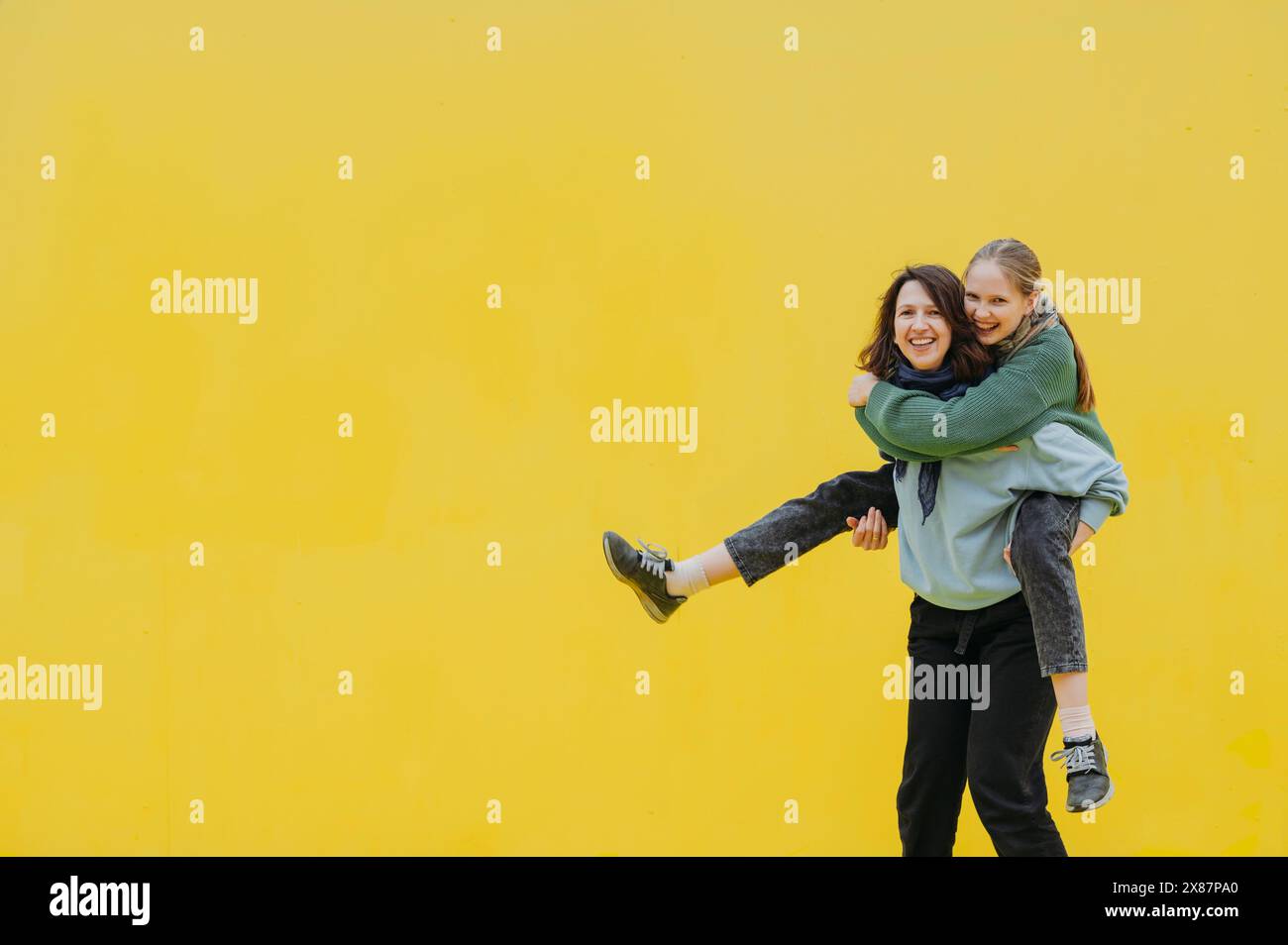 Happy woman giving piggyback ride to daughter against yellow background ...