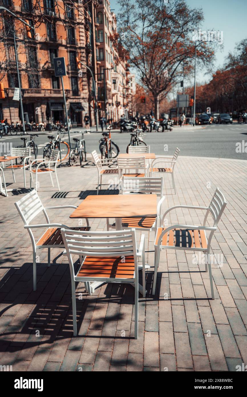 A vertical shot of a sunlit outdoor cafe setting in a bustling Barcelona city with empty tables ...