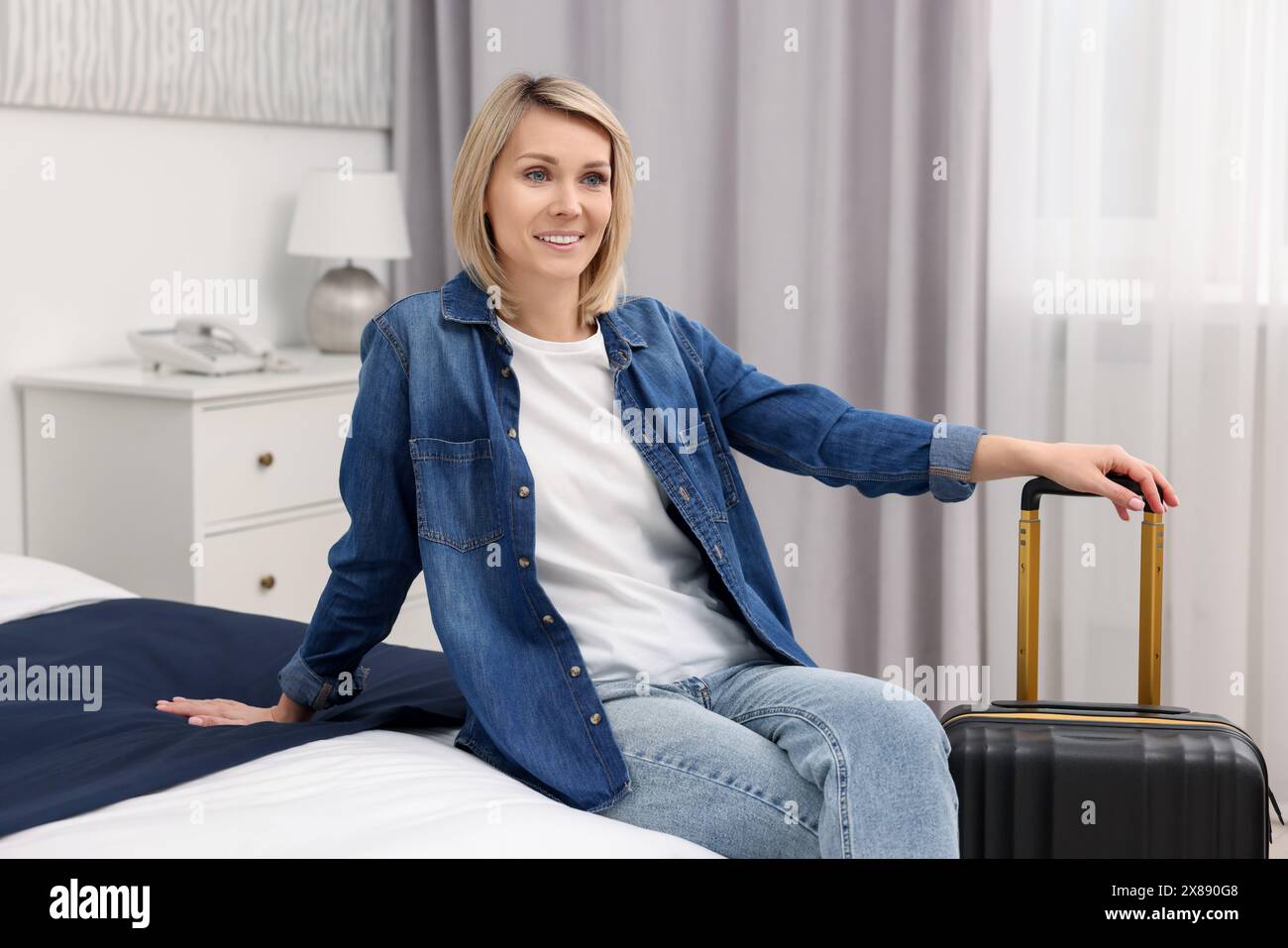 Smiling guest with suitcase relaxing on bed in stylish hotel room Stock ...