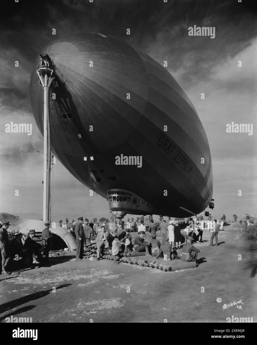 The Graf Zeppelin on the ground in a tied down position with the crowd of people sitting and ...