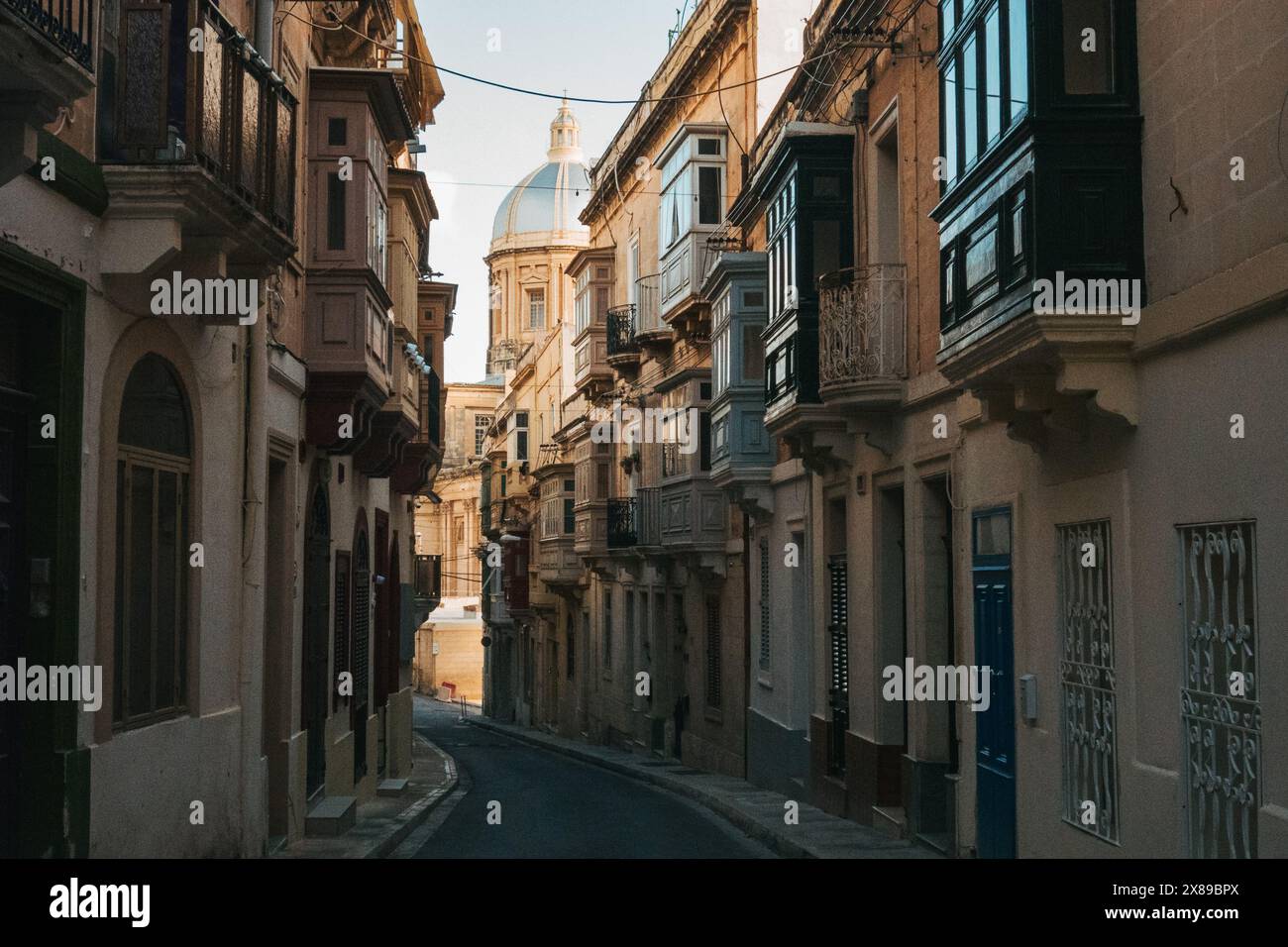 A narrow street in Valletta, Malta, in shadow, with a church dome in ...