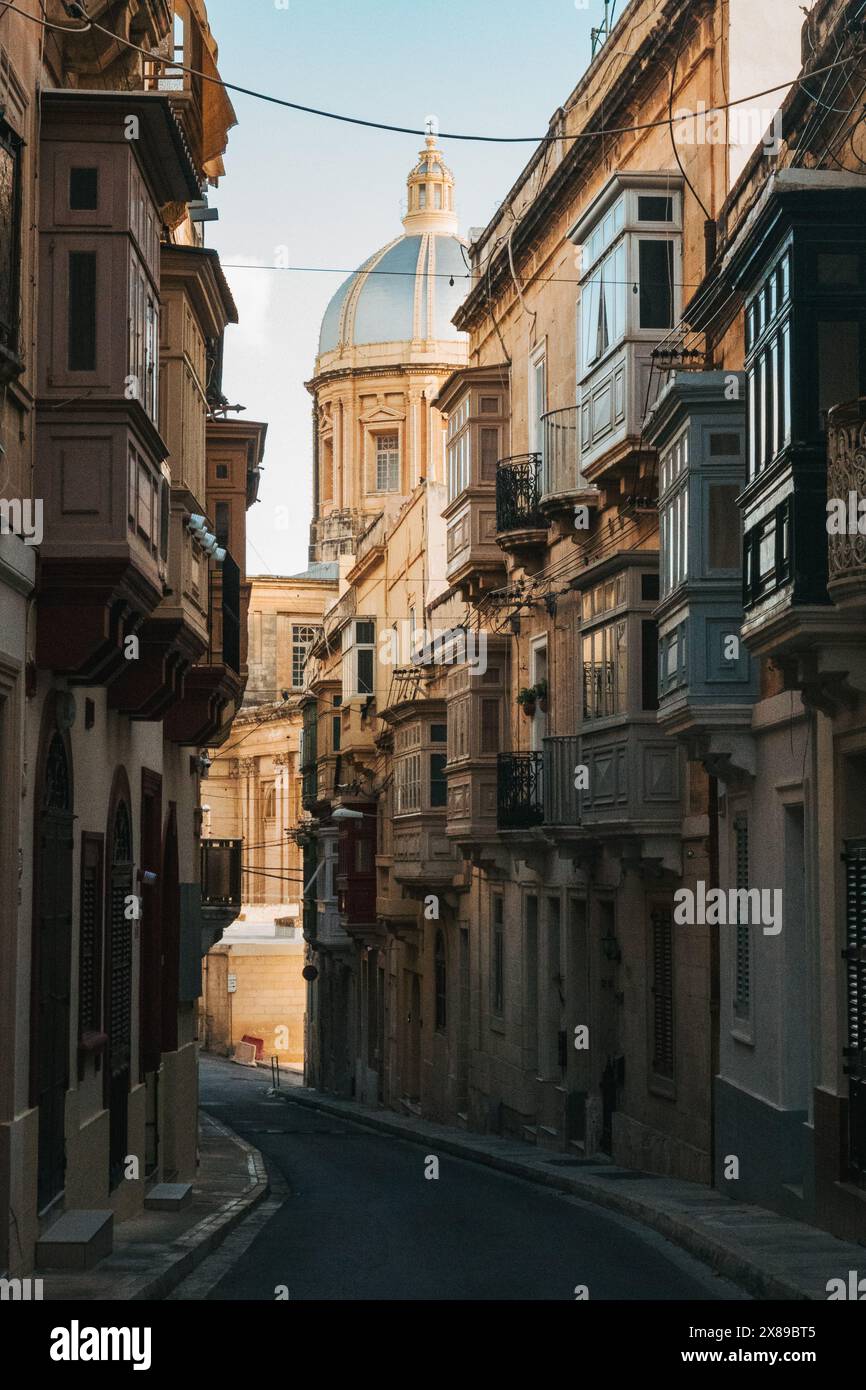 A narrow street in Valletta, Malta, in shadow, with a church dome in ...