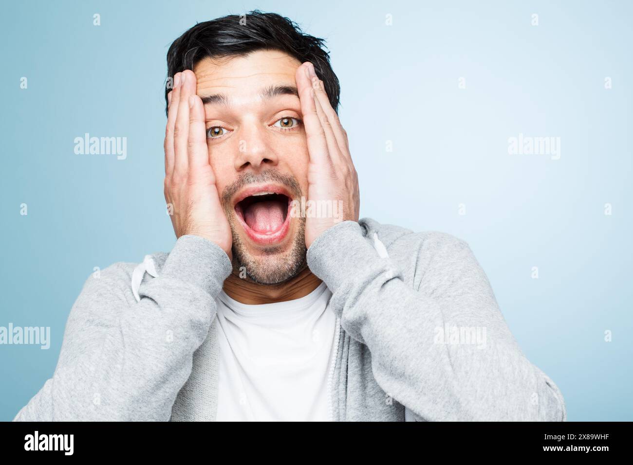 Man, shocked or surprised in portrait with hands on face in blue studio ...