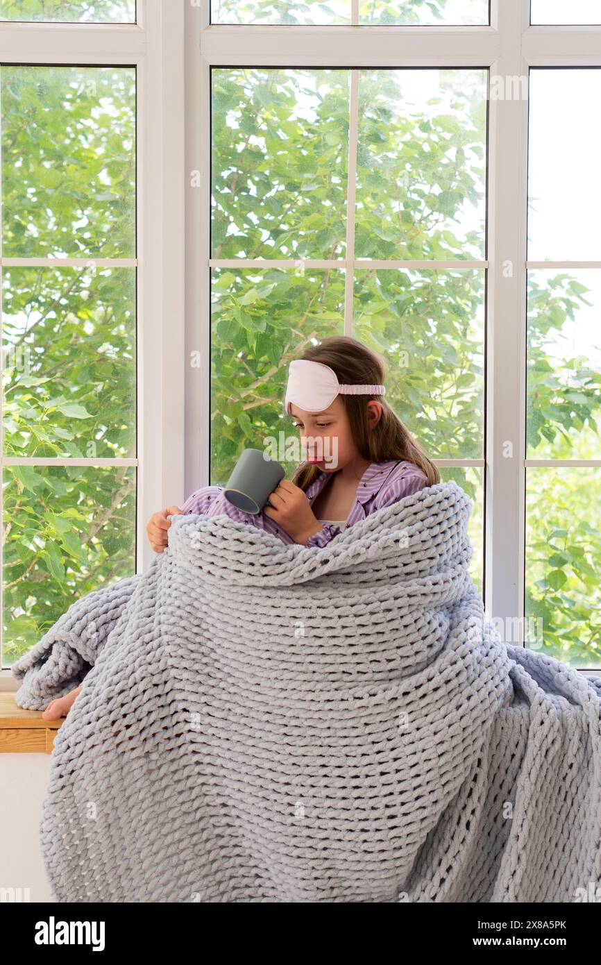 Teenage girl sipping from a mug, cozily wrapped in a grey knitted ...
