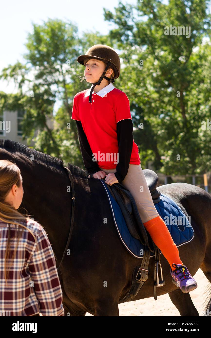 Young rider girl on black horse receiving instruction in sunlight ...