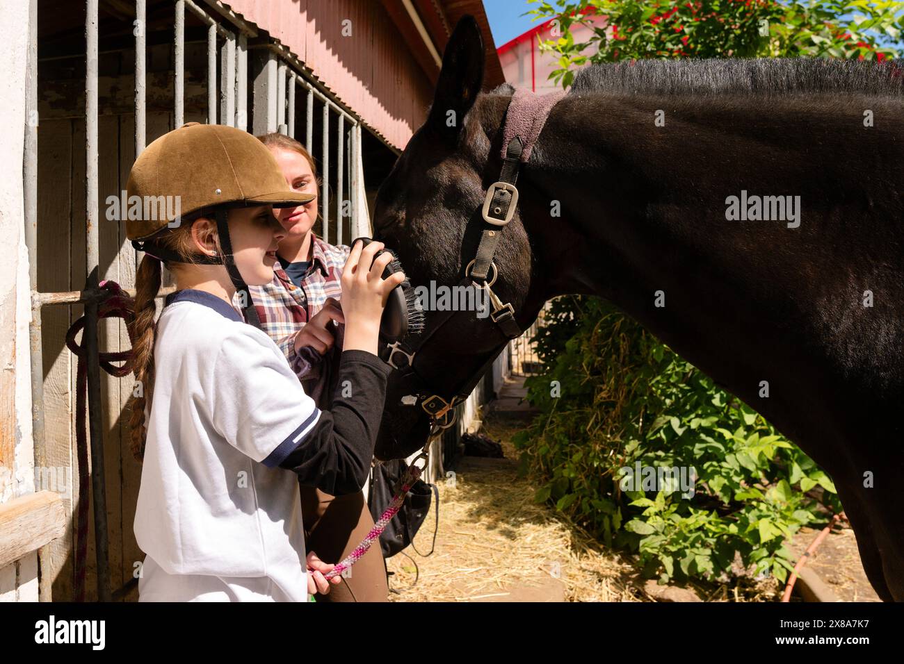 A young girl in a helmet brushes a horse's nose while an instructor ...