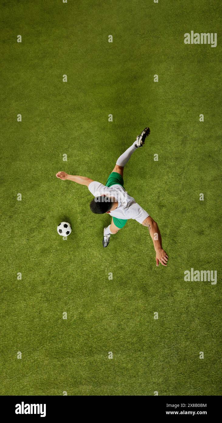 Aerial view of young man, soccer player in green shorts and white jersey striking soccer ball on ...