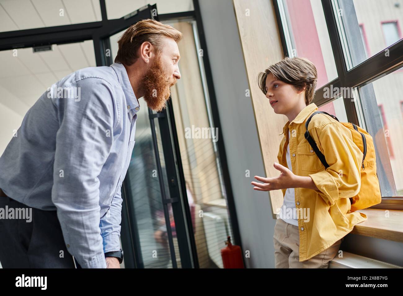 A man stands beside a little boy in front of a window, engaging in a ...