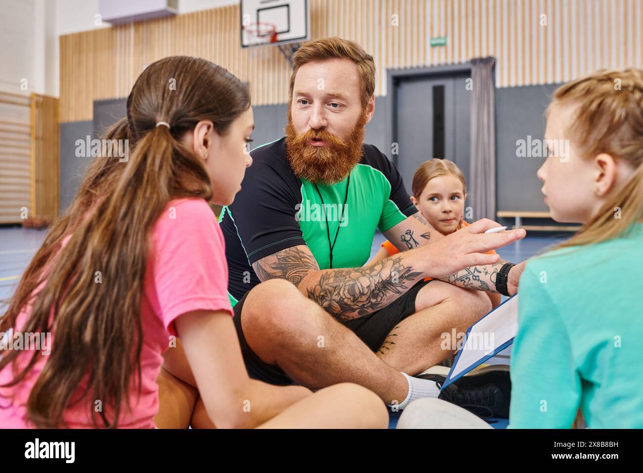 A bearded man sits on a basketball court surrounded by diverse children ...