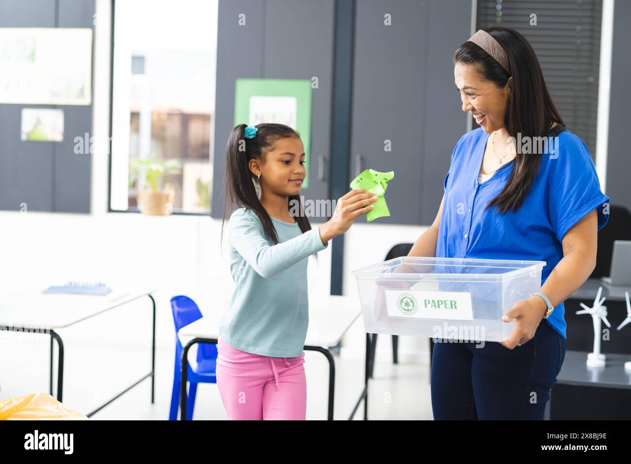 Biracial middle-aged teacher and young girl recycle paper in school ...