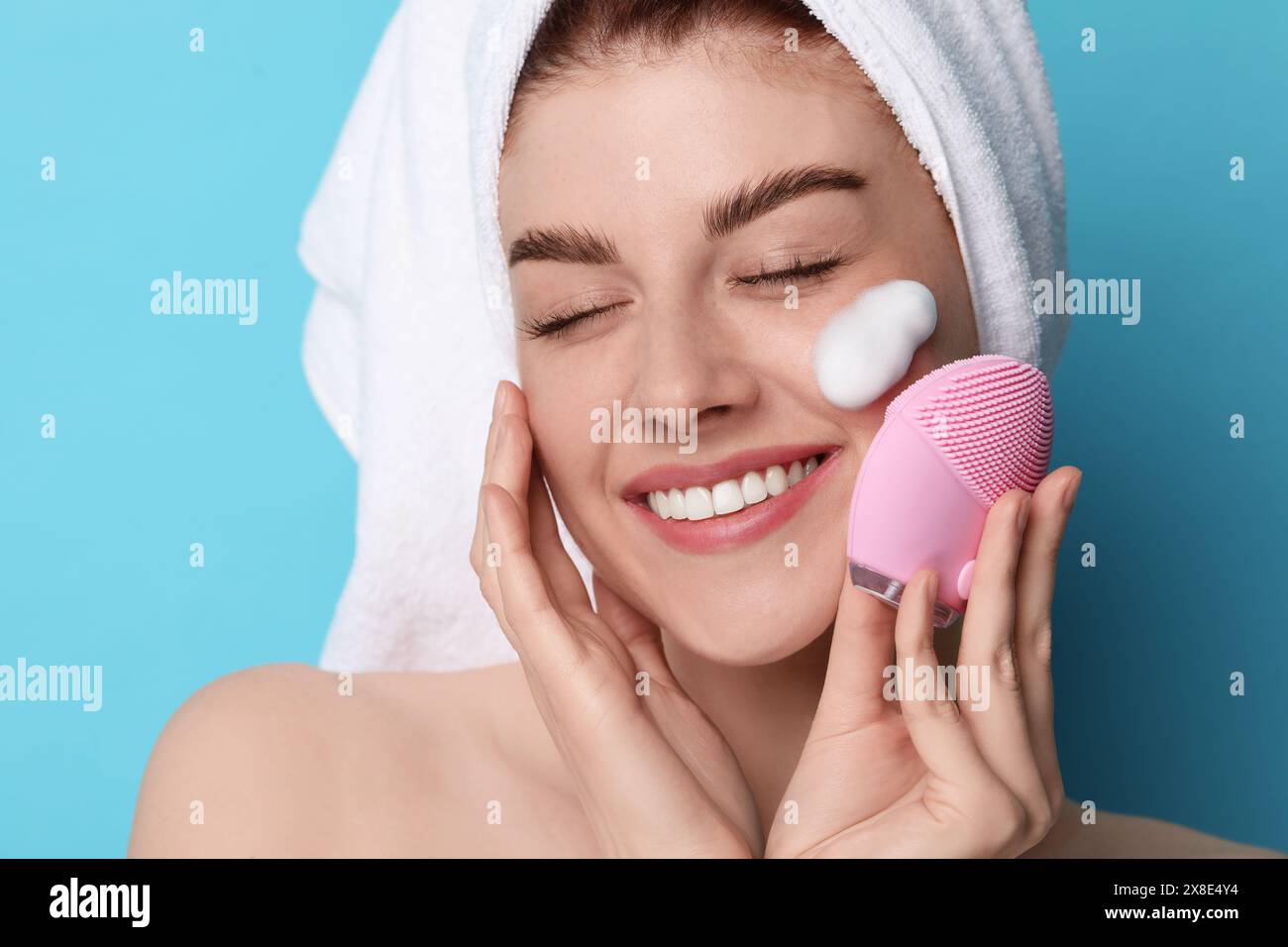 Young woman washing face with brush and cleansing foam on light blue ...