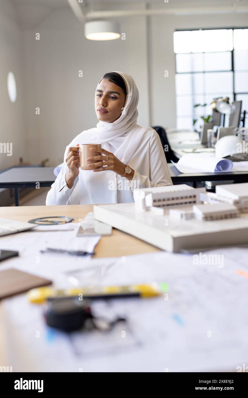 At modern office, Indian woman in white hijab and blouse holding mug ...