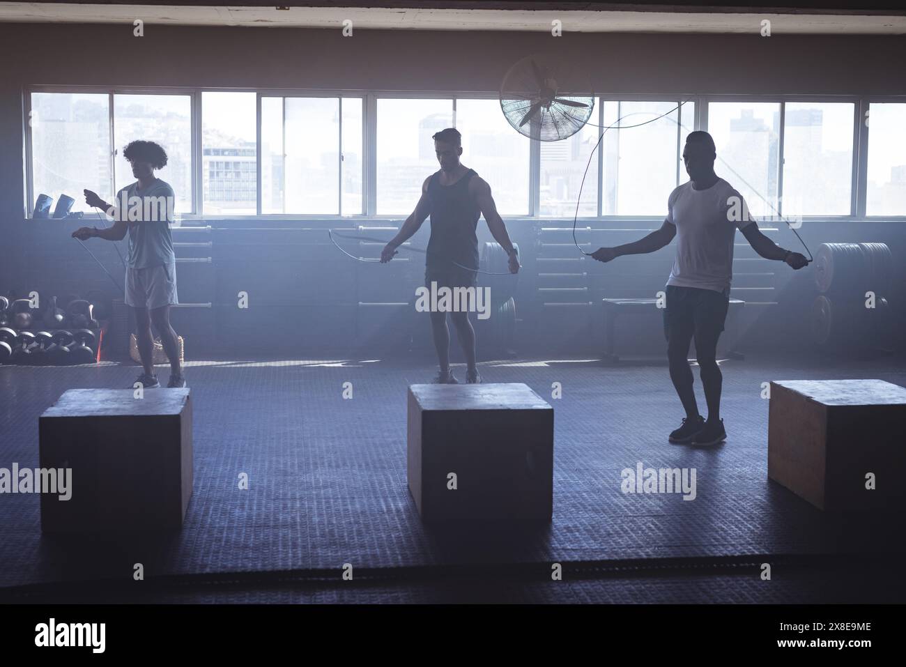 At gym, three fit young diverse men jump roping with large windows in ...