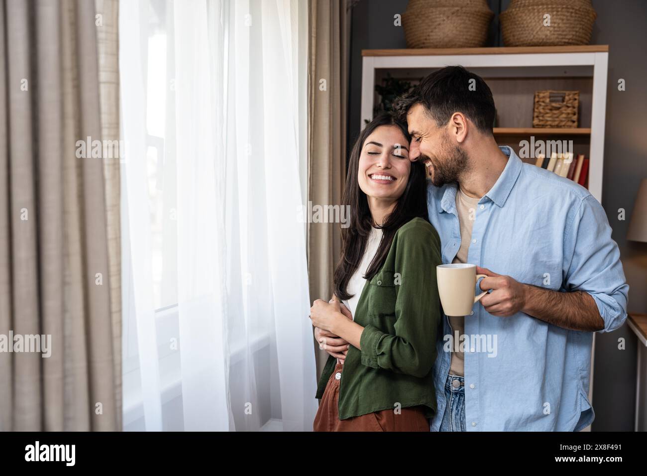 Young happy couple standing near window in their new home looking out ...