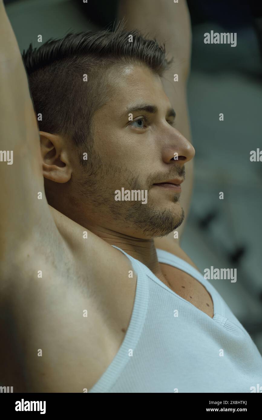 man, dressed in a white tank top, stretches his arms overhead in a gym ...