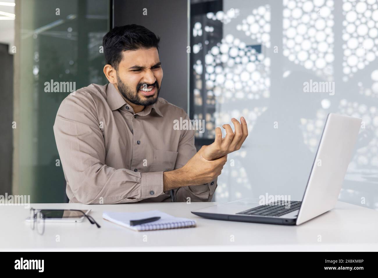 Amid a modern office setup, a businessman grimaces in pain, grimacing, while working on a laptop ...