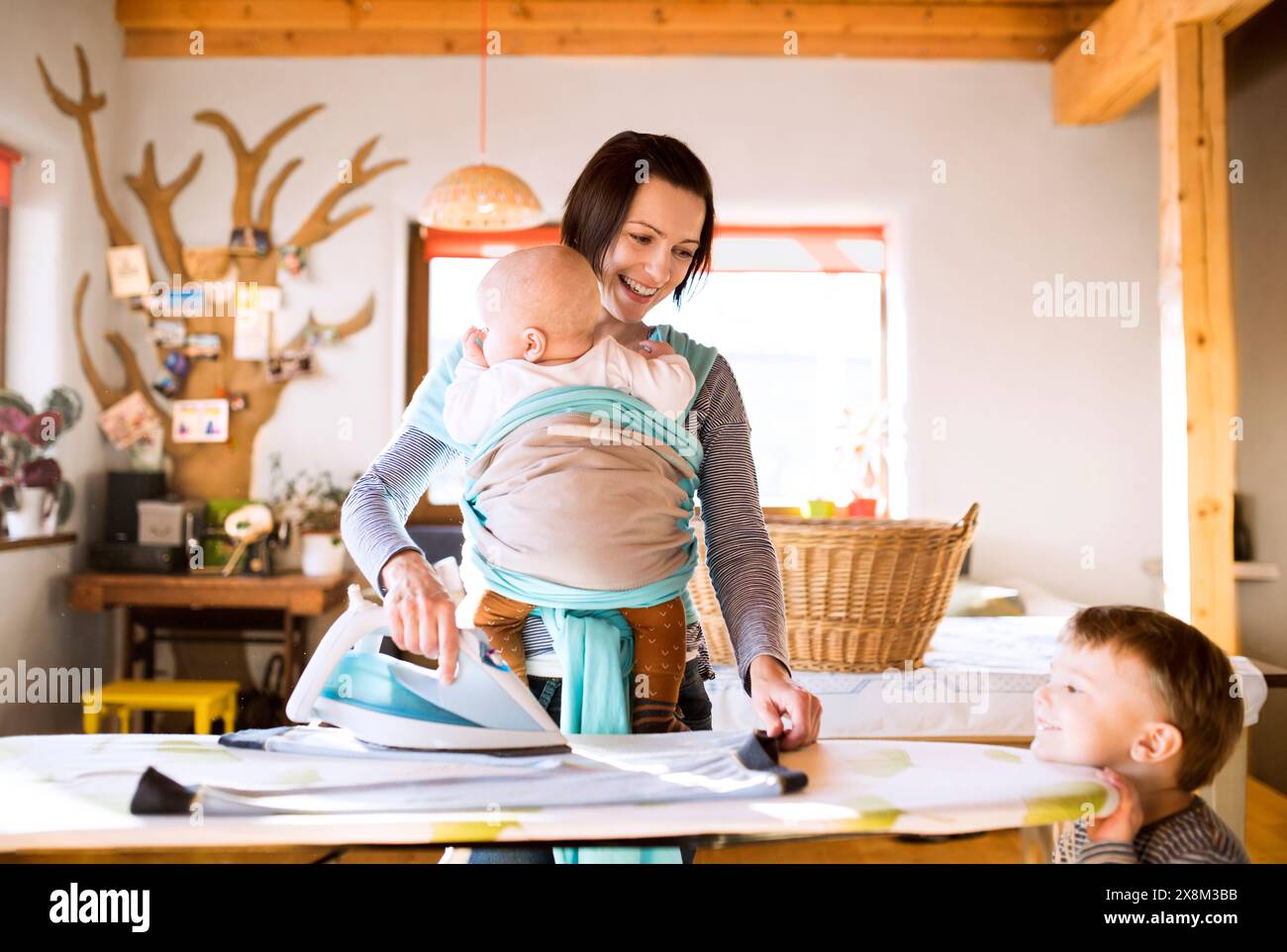 Mother carrying small baby in wrap, older son helping with ironing ...