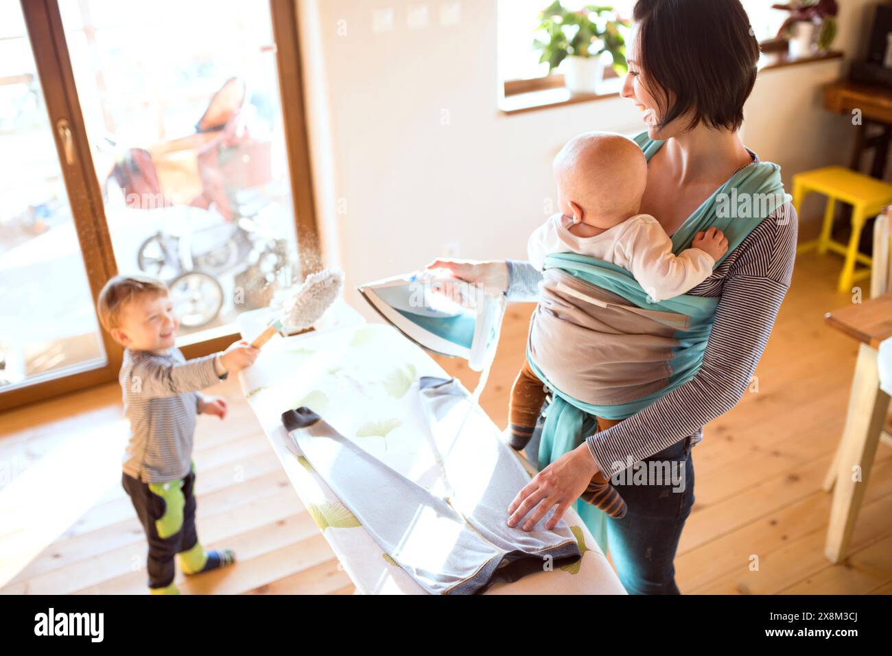Mother carrying small baby in wrap, older son helping with celaning ...