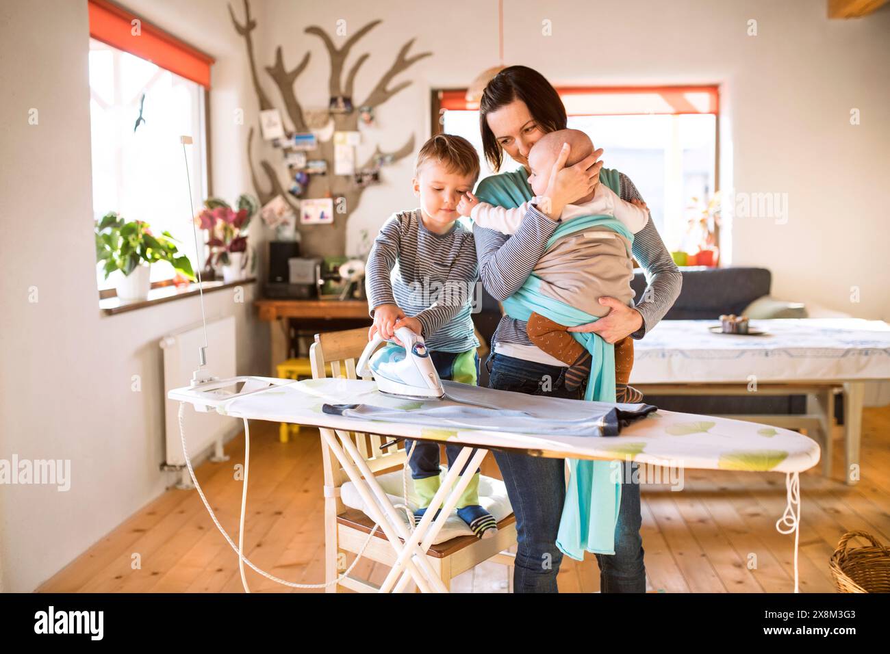 Mother carrying small baby in wrap, older son helping with ironing ...