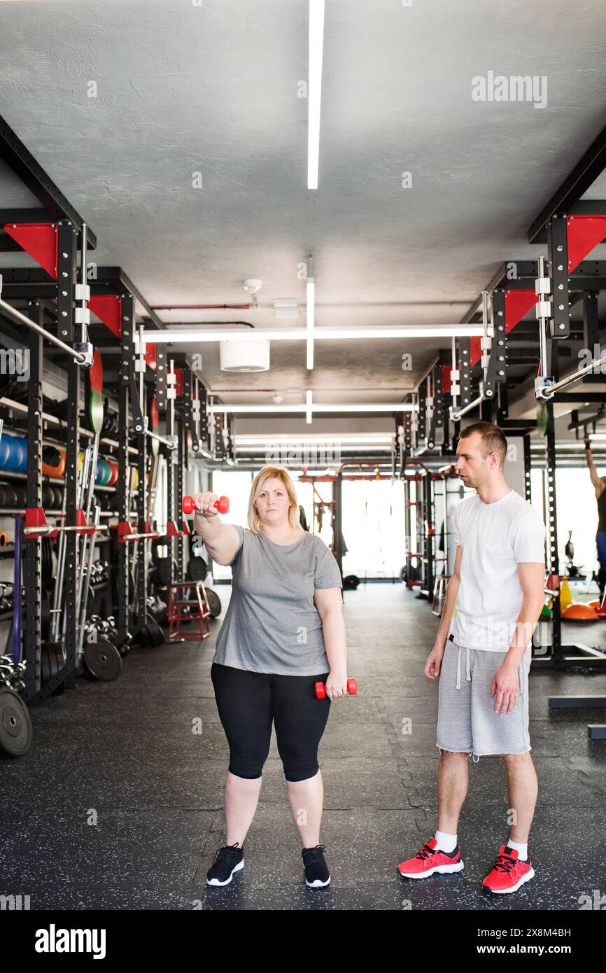 Overweight woman exercising in gym, using dumbbells. Personal trainer ...